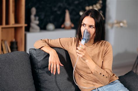 흡입기를 가진 불행한 아픈 백인 젊은 갈색 머리 여성 기침과 독감 치료를 위해 분무기와 흡입기를 사용하여 흡입을하고 소파에 집에 앉아있다 지친 소녀는 휴식과 치료가