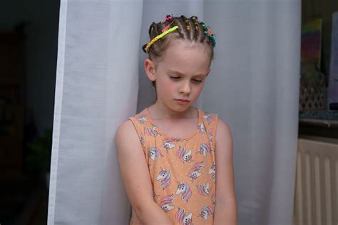 형형색색의 작은 땋은 머리를 한 작고 귀엽고 슬픈 소녀가 방 입구의 하얀 커튼을 배경으로 서 있습니다 어린아이와 같은 슬픔과