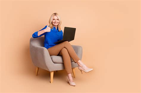 캐주얼 한 파란색 셔츠를 입고 웃는 성숙한 여성이 베이지 색 배경의 안락 의자에 앉아서 노트북으로 작업하고 엄지 손가락을 치켜 올리는 제스처를 보여줍니다 45 49세에 대한