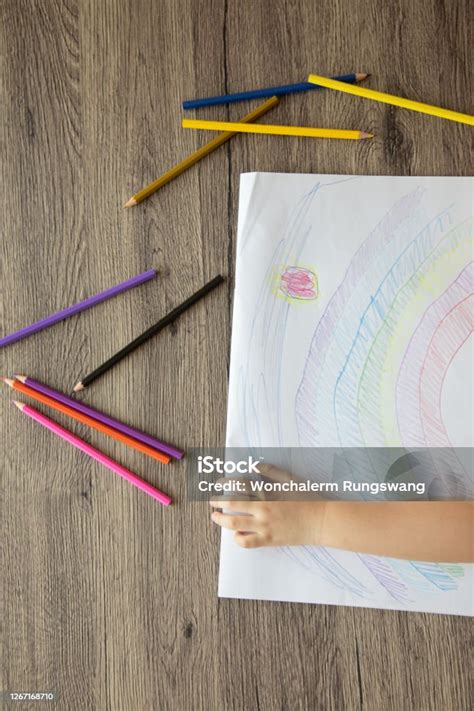 위에서 찍은 사진은 미취학 아동이 나무 테이블에 놓인 흰색 종이에 컬러 연필로 그려진 다채로운 무지개 드로잉을 위해 창의력과 즐거움을 개발하는 법을 배우는 것을 볼 수