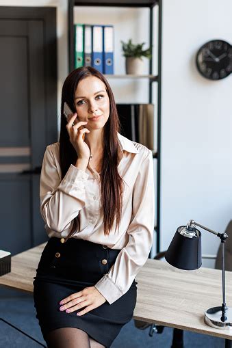 비즈니스 토크 쾌활한 젊은 아름다운 여자는 휴대 전화로 이야기하고 그녀의 작업장에서 테이블에 기대고있는 동안 창문을 바라 봅니다 사무실에 대한 스톡 사진 및 기타 이미지