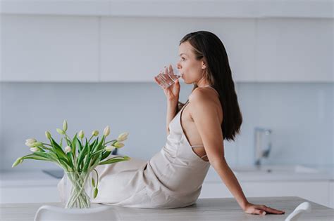 이브닝 드레스를 입은 사려 깊은 스페인 젊은 여성이 튤립으로 가득 찬 꽃병이 물을 마시며 식탁에 앉아 있습니다 긴 머리를 가진 아름다운 백인 여성은 집에서 하루를 시작합니다