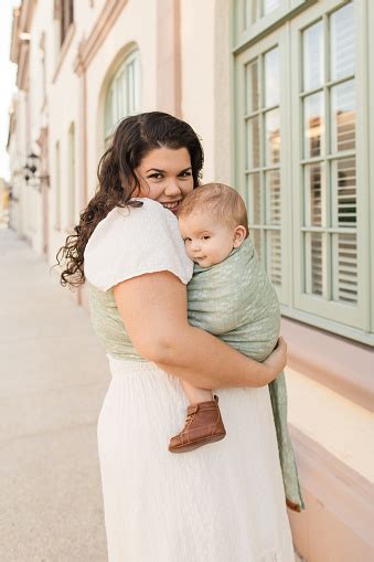30 세의 쿠바 어머니와 5 개월 된 쿠바 계 미국인 아들과 현자 녹색 반지 슬링 베이비 캐리어를 타고 2022 년 봄에 플로리다 팜 비치 의 우아한 보도를 따라 걷고