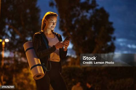 환한 미소를 짓고 있는 자신감 넘치는 백인 젊은 여성이 밤에 공원에서 요가 수업을 마치고 친구를 기다리는 스마트폰을 사용하여