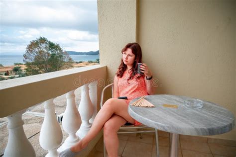 Young Woman In Bright Dress Is Sitting On Chair On Hotel Balcony