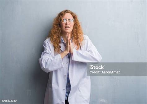 빨강 머리 젊은 의사 여자 회색 그런 지 벽 구걸 코트를 착용 하 고 손으로 얼굴 매우 감정적이 고 걱정에 희망 식 함께 기도 통해 용 서에 대 한 요구 종교 개념입니다