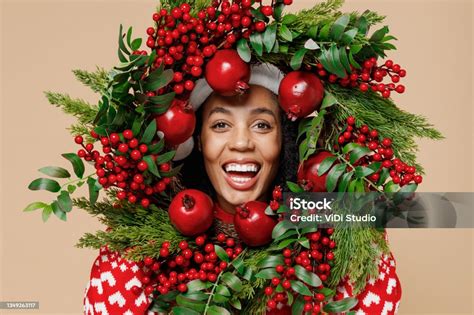 크리스마스 모자 스웨터를 입은 젊은 아프리카계 미국인 산타 여성이 파스텔 베이지 색 배경 스튜디오 초상화에 고립 된 축제 화환을 통해 모습을 유지합니다 새해 복 많은 설 날