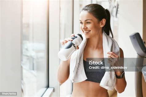휴식 시간 체육관에서 창 앞에 서 있는 동안 그녀의 어깨에 물을 마시는 흰색 수건을 가진 스포츠웨어에서 젊고 쾌활 한 여자 체육관에 대한 스톡 사진 및 기타 이미지 Istock