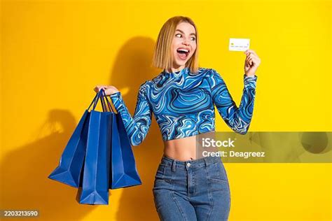 밝은 노란색 배경을 배경으로 파란색 쇼핑백과 기프트 카드를 들고 포즈를 취하며 행복과 흥분을 표현하는 쾌활한 젊은 여성 20 24세에 대한 스톡 사진 및 기타 이미지