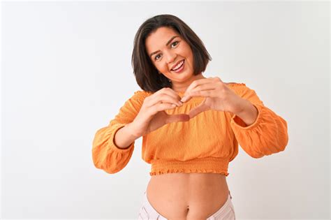 고립 된 흰색 배경 위에 서있는 오렌지 색 티셔츠를 입은 젊은 아름다운 여성은 손으로 심장 기호와 모양을 보여주는 사랑에 미소