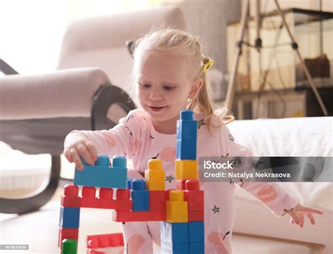 웃고 있는 어린 아이 화려한 큐브를 가지고 노는 금발의 미취학 아동 분홍색 잠옷을 입고 바닥에 앉아 집에 큰 창문이 있는 햇볕이 잘 드는 방 아이들의 초기 발달 2 3 살에