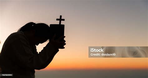 무릎을 꿇고 기도하고 일몰 배경에서 하나님을 경배하기 위해 기독교 십자가를 들고 있는 젊은 여성의 실루엣 기독교인이 무릎을 꿇고 하나님께 기도하는 개념 개념에 대한 스톡