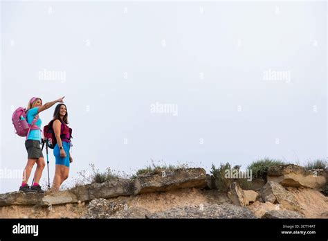 Female Hikers Standing On Cliff Edge As One Points Toward Horizon