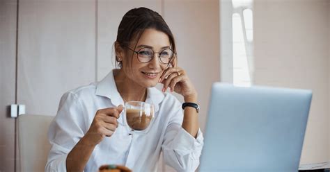 뜨거운 음료 한 잔을 들고 사무실에서 현대적인 노트북을 들고 테이블에 앉아 웃고 있는 아름다운 여자 사진 Unsplash의 아름다운 사람들 이미지