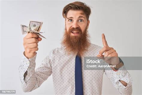 젊은 빨간 머리 아일랜드어 사업가 는 행복한 얼굴 번호 하나 와 손가락을 가리키는 아이디어 또는 질문으로 놀란 고립 된 흰색 배경 위에 서 있는 달러를 들고 가리키기에 대한