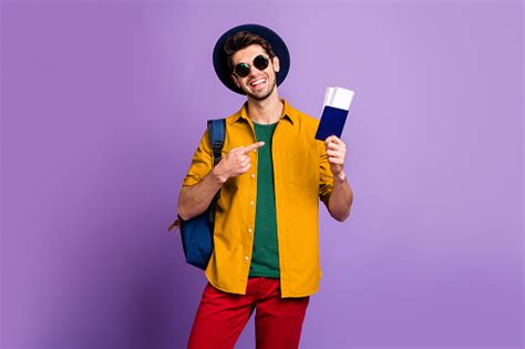 벨벳 모자 배낭 홀드 티켓 공기 비행 포인트 집게 손가락 조언 퍼스트 클래스 착용 노란색 셔츠 빨간 바지 바지 고립 된 보라색 색상 배경을 가진 긍정적 인 관광 남자 보여