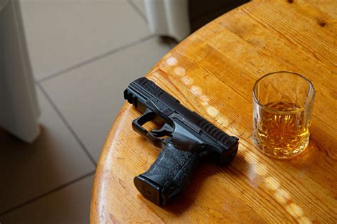 나무 테이블에 검은 손총이 열리고 흐릿한 문이 바닥에 드리워진 그림자가 배경에 있습니다 위스키가있는 유리가 무기 근처에 머물러 있습니다 0명에 대한 스톡 사진 및 기타