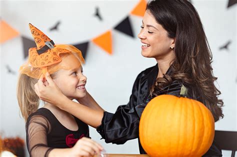 한 어머니가 딸이 귀여운 할로윈 마녀 모자를 고쳐 쓰는 것을 도와주고 둘은 서로를 향해 미소를 짓는다 테이블 위에는 조각된 호박이 놓여 있고 배경에는 축제 분위기의 할로윈
