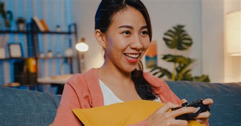 무선 컨트롤러를 사용하는 젊은 아시아 여성은 집에서 밤에 거실의 소파에서 재미있는 행복한 순간을 보내는 비디오 게임을 합니다