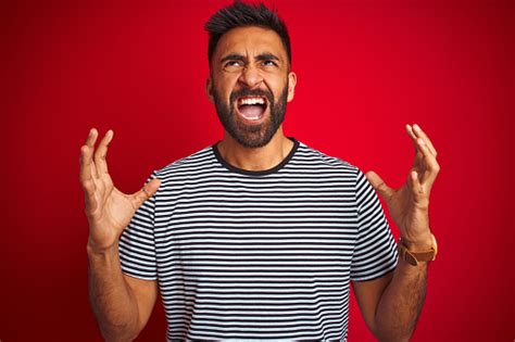 해군 줄무늬 티셔츠를 입은 젊은 잘생긴 인도 남자는 고립 된 빨간 배경에 미친 미친 소리와 공격적인 표정과 팔을 들고 고함 좌절 개념입니다 T 셔츠에 대한 스톡 사진 및