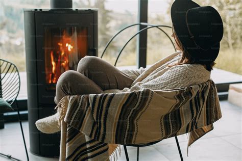 니트 스웨터와 모자를 쓴 세련된 여성이 산의 전망을 감상할 수 있는 현대적인 검은색 벽난로에서 워밍업을 하고 있다 추운 계절에