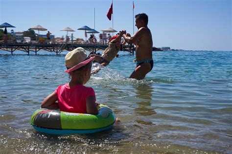 밀짚모자를 쓴 소녀가 해안의 바다에 앉아 가족들이 바다에서 즐거운 시간을 보내는 것을 지켜보고 있다 바닷가에서 가족 휴가를 보내세요 여름 방학 바다 휴일 여름 라이프 스타일