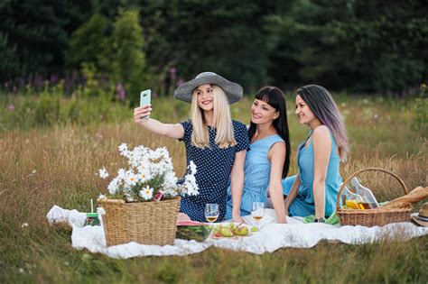 파란 드레스와 모자를 쓴 세 명의 청녀가 격자무늬에 앉아 스마트폰으로 사진을 찍고 있다 숲의 잔디에 야외 피크닉 피크닉 바구니와 와인에 맛있는 음식 3 명에 대한 스톡 사진