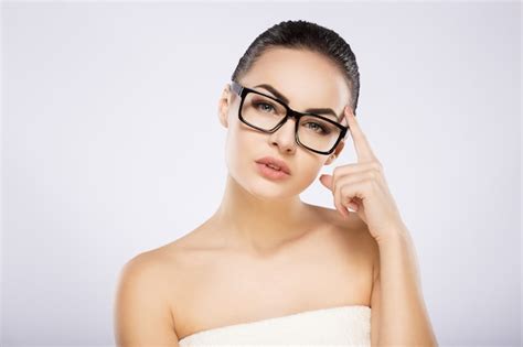 검은 머리가 뒤에 고정 된 화려한 소녀 큰 눈 두꺼운 눈썹 및 안경을 쓰고 회색 스튜디오에서 포즈를 취하는 벌거 벗은 어깨 프리미엄 사진