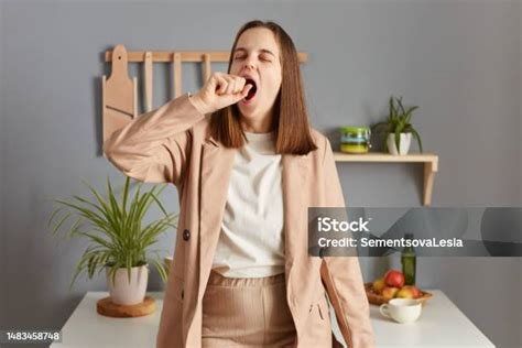 졸린 노동자 일찍 깨어났다 베이지색 재킷을 입은 갈색 머리의 하품하는 여자는 손바닥으로 입을 가리고 집 부엌 인테리어에 서서 잠을 이루지 못한다 30 34세에 대한 스톡