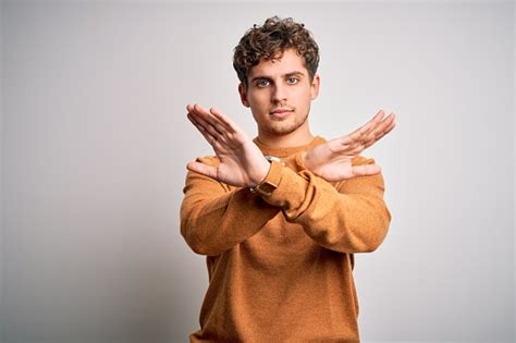 젊은 금발의 잘생긴 남자 와 곱슬 머리 입고 캐주얼 스웨터 위에 흰색 배경 거부 표현 횡단 팔 을 하고 부정적인 기호 화가 얼굴