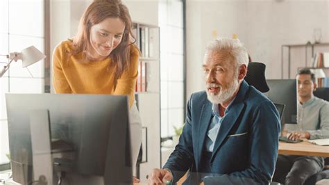 데스크톱 컴퓨터에서 작업 하는 책상에 앉아 잘 생긴 중년 창조적 인 엔지니어는 아름 다운 여성 프로젝트 관리자와 토론을 친절한 동료채팅 솔루션 찾기와 함께하는 사무실 2명에