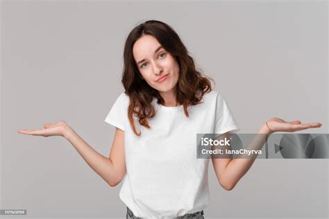 예쁜 갈색 머리 여자 에 A 흰색 티 어깨를 으쓱 그녀의 어깨 Beacause 그녀는 회색 배경 위에 고립 된 분개 서 광고 의 공간을 복사합니다 T 셔츠에 대한 스톡 사진
