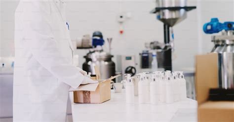 판지 상자에 로션이 든 병을 포장하는 멸균 옷을 입은 남자의 그림 밝은 실험실에 서서 제품을 포장합니다 사진 Unsplash의 개념 이미지