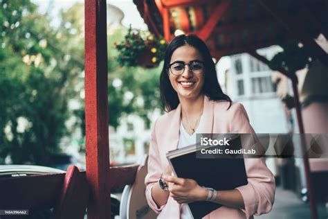 도시 환경에서 포즈를 취하는 스케치북을 든 쾌활한 여성 저널리스트의 절반 길이 초상화 도시에서 여가 시간 동안 카메라를 향해 웃고 있는 눈 교정을 위한 고전적인 안경을 쓴