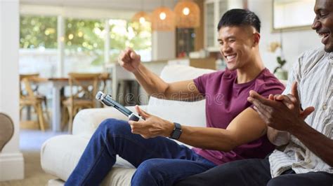 Same Sex Male Couple Or Friends On Sofa At Home With Handheld Mobile Same Sex Male Couple Or Friends On Sofa At Home With Handheld Mobile