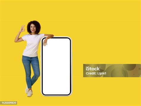 Gesturing Ok 젊은 아프리카 계 미국인 여성 행복은 흰색 티셔츠와 노란색 배경에 고립 된 데님 청바지를 입은 흰색 화면이있는 크고 거대한 스마트 폰에 기대어