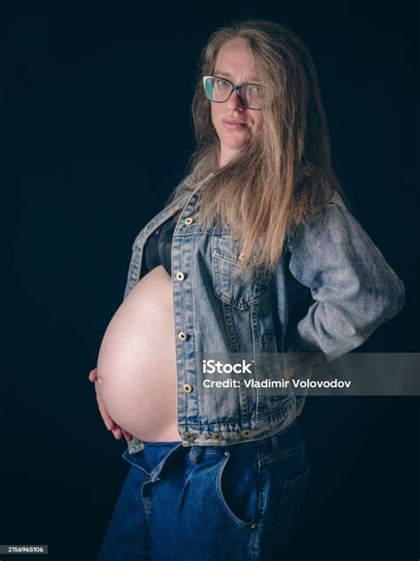 A Pregnant Woman Wearing Glasses Stands Sideways Against A Black