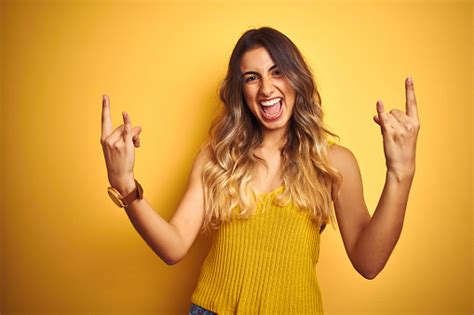 노란색 고립 된 배경 위에 티셔츠를 입고 젊은 아름다운 여성은 손을 위로 바위 기호를하고 미친 표정으로 외칩니다 음악 스타 무거운 음악 개념 T 셔츠에 대한 스톡 사진 및