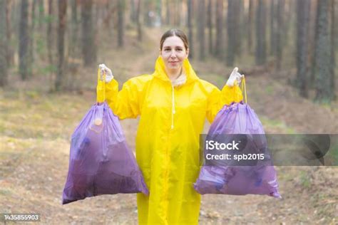 노란색 비옷을 입은 웃는 여성 자원 봉사자 보호론자가 보라색 쓰레기 봉투 두 개를 들고 있습니다 자연 보호에 대한 그의 업적을 보여줍니다 가비지 수집 소나무 숲을 배경으로