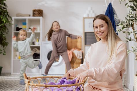 세탁기 옆의 배경에서 세탁을 위해 옷을 분류하는 바닥에 앉아있는 웃는 여성의 초상화 두 귀여운 어린 소녀가 춤을 추고 속이고 함께 시간을 보내고 있습니다 2명에 대한 스톡