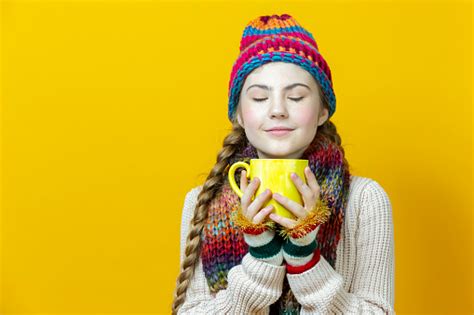 따뜻한 계절 모자와 니트 스카프를 들고 노란색 배경에 차 또는 커피를 마시는 동안 큰 노란색 컵을 들고있는 Winsome Calm Caucasian Teenage Girl