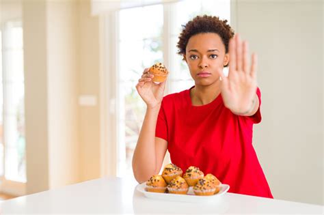 진지하고 자신감있는 표정 방어 제스처로 정지 표지판을하는 오픈 손으로 초콜릿 칩 머핀을 먹는 젊은 아프리카 계 미국인 여성 가정 생활에 대한 스톡 사진 및 기타 이미지