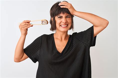 머리에 손으로 스트레스를 받은 고립된 흰색 배경 위에 서 있는 젓가락을 사용하여 초밥을 먹는 젊은 아름다운 여성은 수치심과 놀라움의 얼굴 분노와 좌절에 충격을 받았습니다