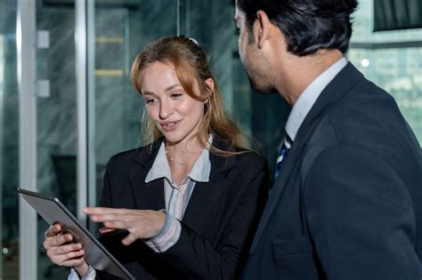 백인 여성 직원이 사무실 협업 중 중동 남성 동료에게 디지털 태블릿으로 아이디어를 발표하는 모습 직장에서 공식적인 비즈니스 복장을 입고 미소를 지으며 함께 서 있는 모습