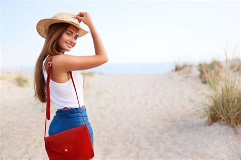 세련 된 여성은 밀 짚 모자 여름 데님 반바지와 빨간색 세련 된 가방에 해변에 산책 매력적인 검게 여자는 바다 해 안에 그녀를 따르도록 초대 합니다 웃는 소녀는 바다로 간다