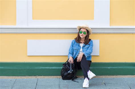 Smath 젊은 아시아 여성 여행자 청바지 재킷 선글라스와 밀짚 모자를 입고 통로 측면에 앉아 노란색 벽 휴식 아시아에서 도시 여행 하는 동안 거리에 대한 스톡 사진 및