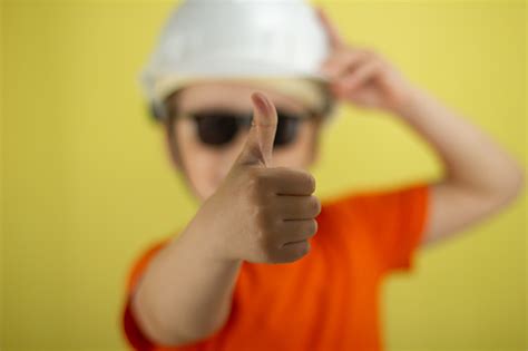 건설 헬멧을 쓴 소년이 엄지손가락을 치켜들고 있다 주황색 바탕에 있는 아이가 엄지손가락으로 교실을 보여주고 있다 선글라스와 건설 헬멧을 쓴 아이 격리 건설 산업에 대한 스톡
