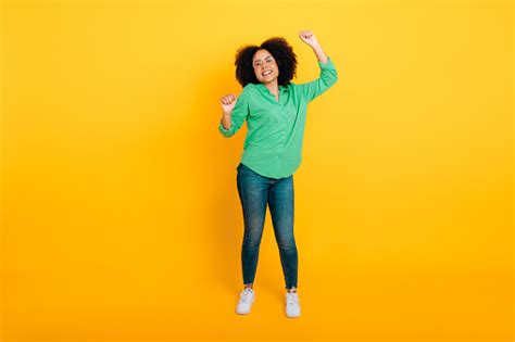 쾌활한 아프리카계 미국인 또는 브라질의 세련된 곱슬머리 여성의 전체 길이 사진 고립된 노란색 배경에 서서 주먹으로 몸짓 웃고 기뻐하고 춤추고 즐거운 감정을 경험합니다 승리