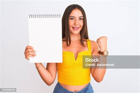 고립된 흰색 배경 위에 서있는 노트북을 들고있는 젊은 아름다운 학생 소녀는 자랑스럽게 비명을 지르며 승리와 성공을 축하하며 매우 흥분하고 환호하는 감정을 갈색 머리에 대한