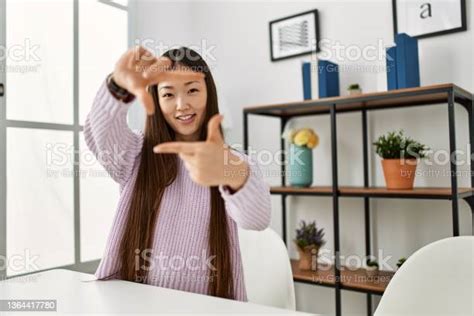집에서 테이블에 앉아 캐주얼 한 옷을 입은 어린 중국 소녀는 행복한 얼굴로 손과 손가락으로 프레임을 만드는 미소를 지었습니다 창의력과 사진 개념 개념에 대한 스톡 사진 및